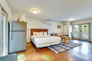 Quarto com cama, cabeceira de madeira, mesinha, geladeira e porta que dá para uma varanda.