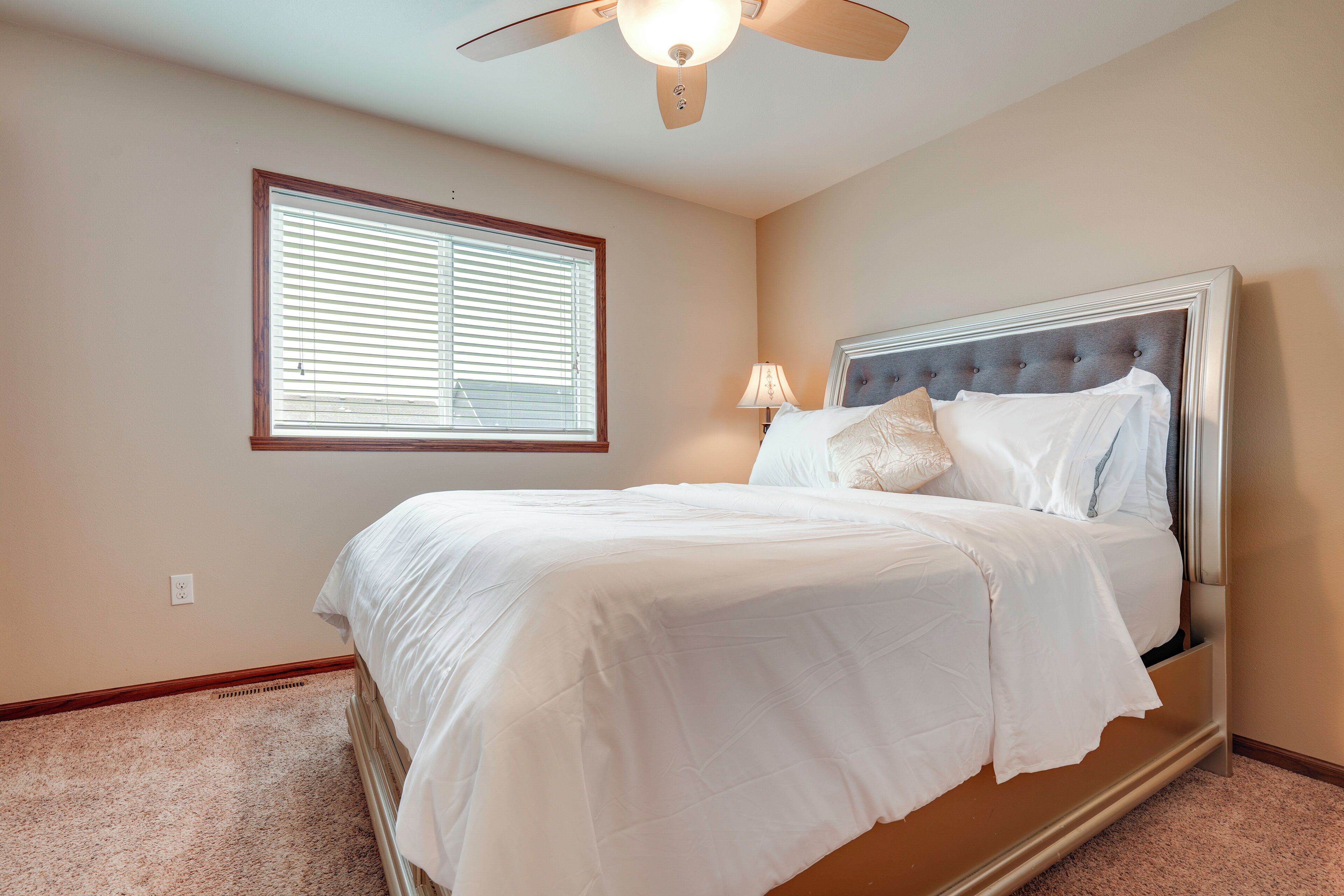 Quarto com cama, ventilador de teto, janela com persianas e mesa de cabeceira com abajur.