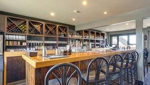 Uma área de bar com balcão de madeira, banquetas e prateleiras cheias de garrafas e copos.