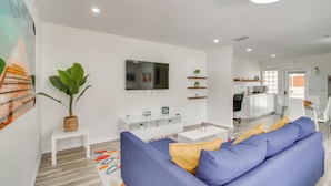 Uma sala de estar moderna com um sofá azul, uma mesa de centro branca e uma televisão de tela plana. Há um pequeno armário branco com objetos decorativos e uma planta em vaso.