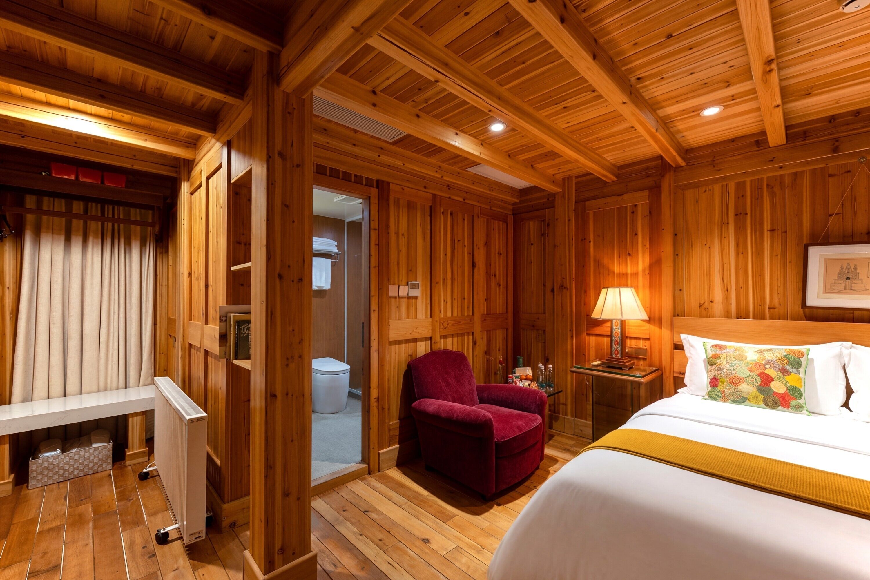 Um quarto com painéis de madeira, uma cama, uma cadeira vermelha, uma mesinha de cabeceira e um radiador.