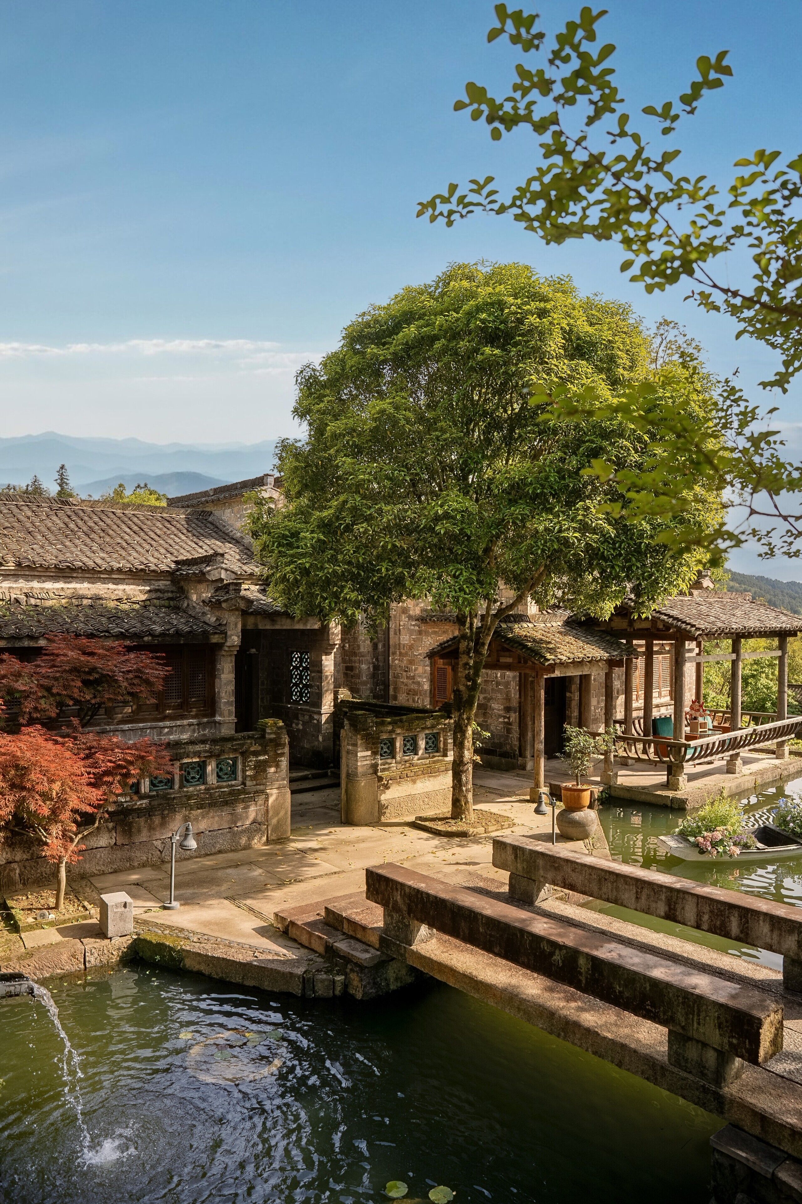 Um pátio tradicional com uma ponte de pedra, uma árvore e um pequeno lago.