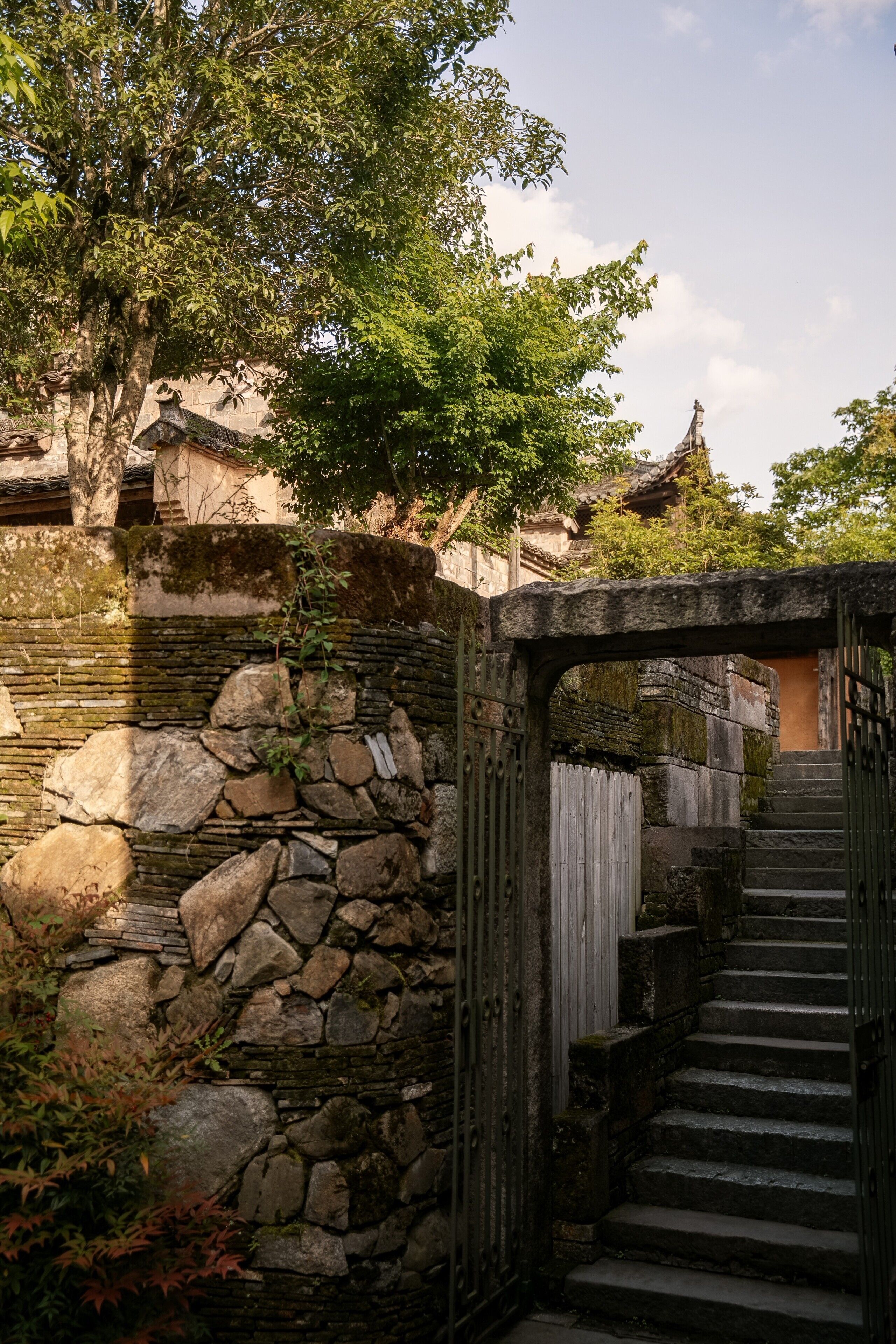 Um muro de pedra com um portão, uma escadaria e um edifício tradicional ao fundo.