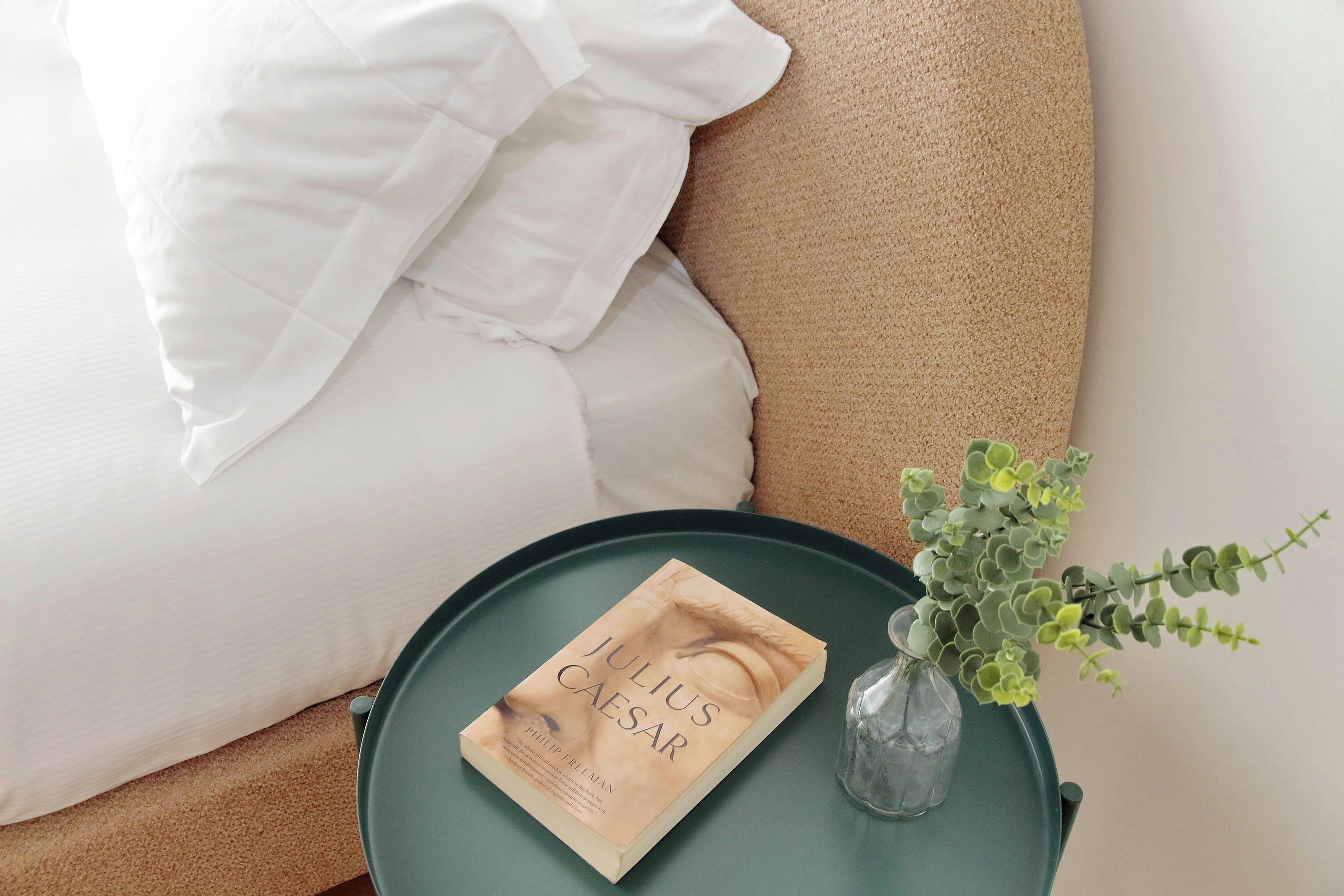 Cama com roupa de cama branca, mesa de cabeceira verde com um livro intitulado 'Julius Caesar' e um vaso com plantas verdes.