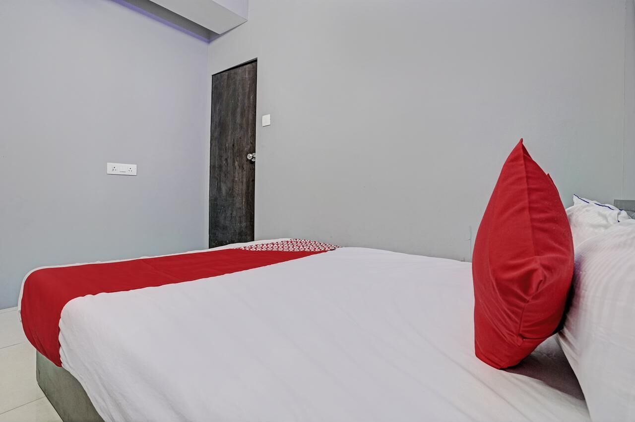 Cama bem arrumada com edredom e almofadas vermelhas e brancas, porta escura e parede clara com tomada.
