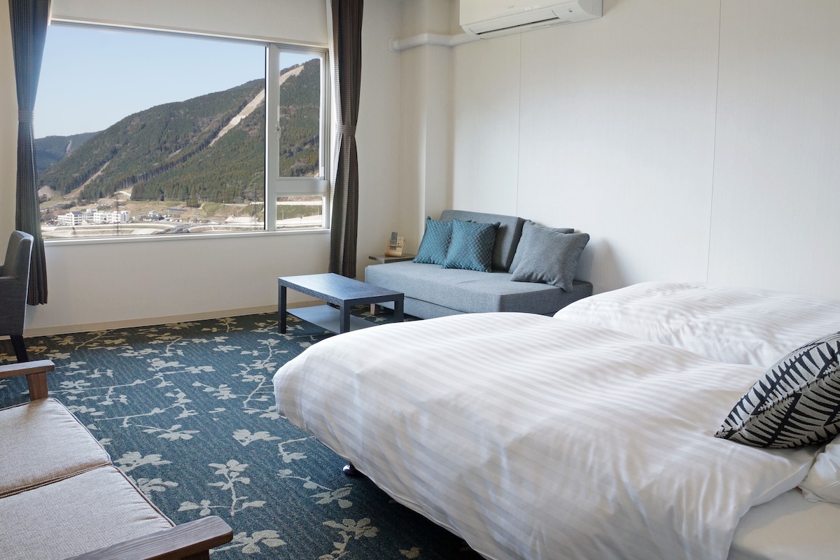 Quarto de hotel com cama, escrivaninha, cadeira, mesinha e sofá. Há uma janela com vista para uma montanha e um edifício.