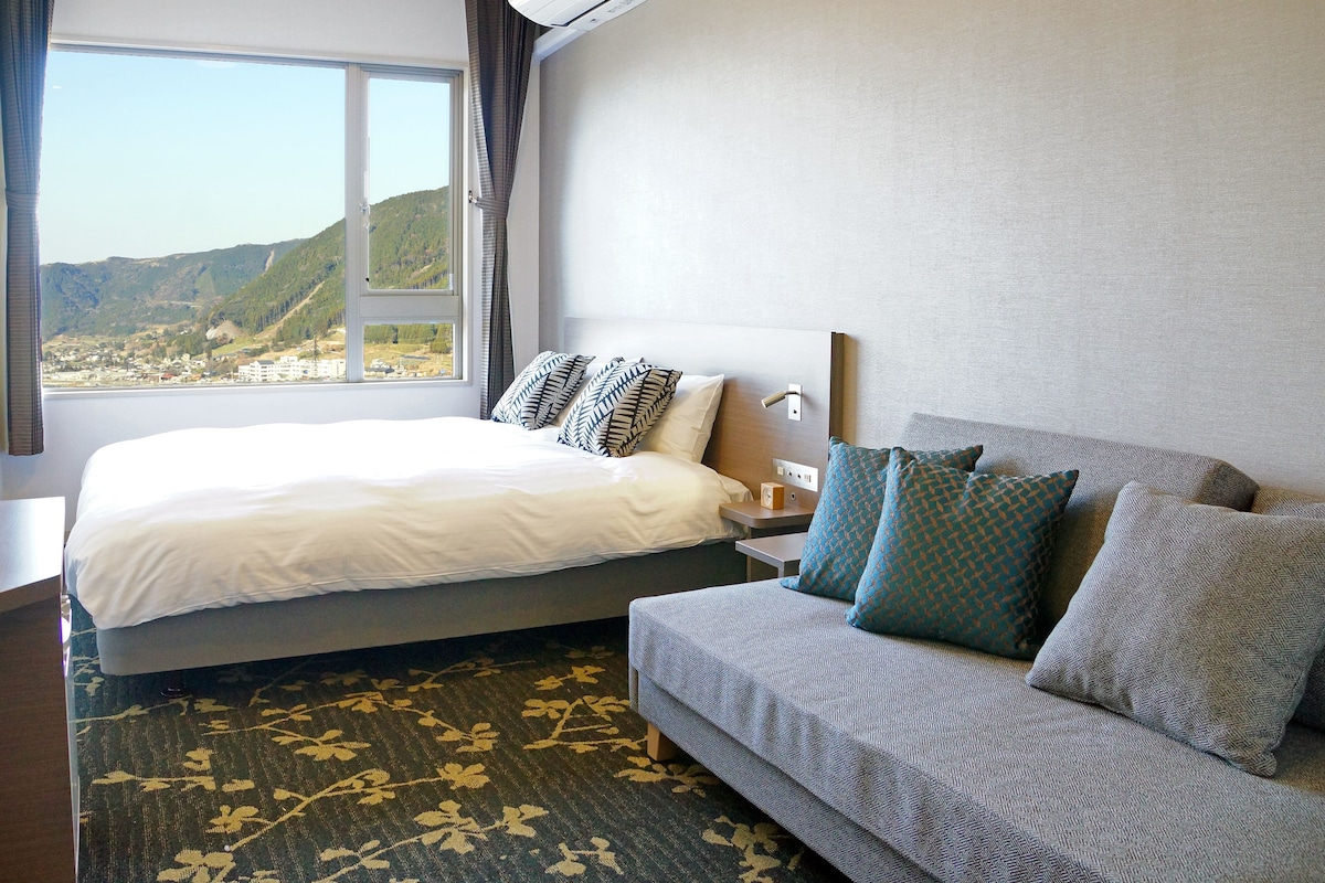 Quarto de hotel com cama, sofá, escrivaninha e vista para uma paisagem montanhosa.