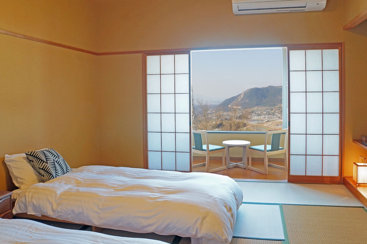 Quarto de hotel com cama, uma pequena mesa com cadeiras, vista para uma paisagem montanhosa através de uma porta deslizante e uma porta deslizante em estilo tradicional japonês.