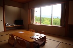 Quarto com piso de tatame, uma mesa de madeira, cadeiras, televisão e vista para o verde através de grandes janelas.