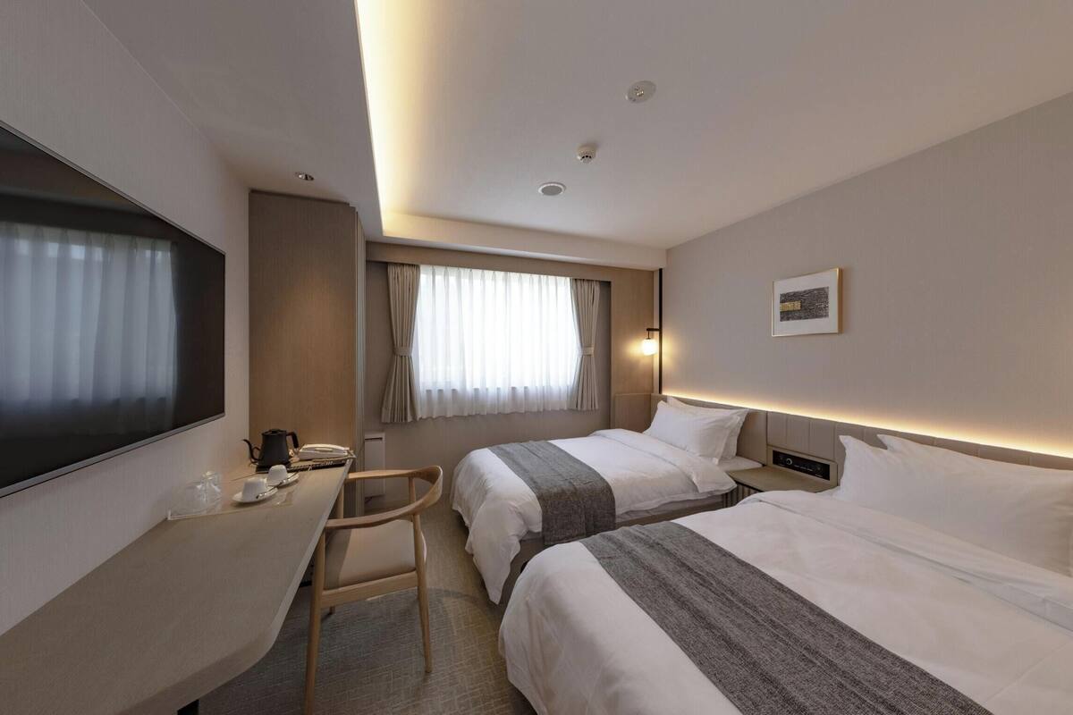 Quarto de hotel com duas camas, uma mesa, uma cadeira, uma televisão e uma janela com cortinas.
