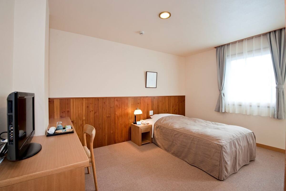 Quarto de hotel com cabeceira de madeira, cama com cobertura bege, escrivaninha com computador, cadeira, mesa de cabeceira com abajur e janela com cortinas.