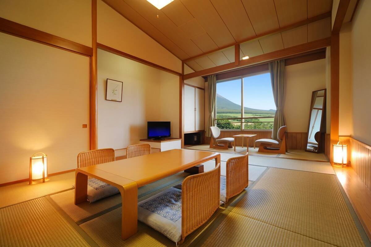 Um quarto japonês tradicional com tatames, uma mesa baixa, cadeiras, televisão e vista para o exterior.