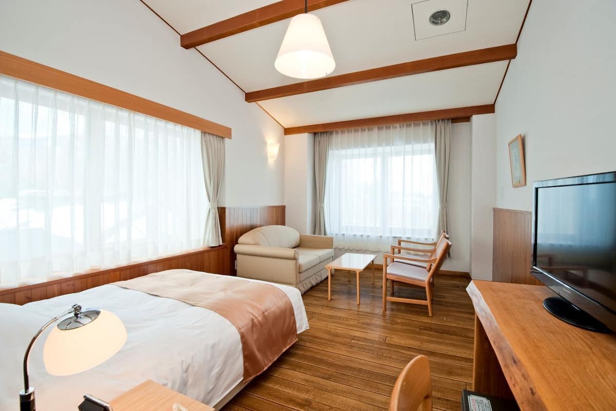 Quarto de hotel com cama, escrivaninha, cadeira, televisão e janela com cortinas.