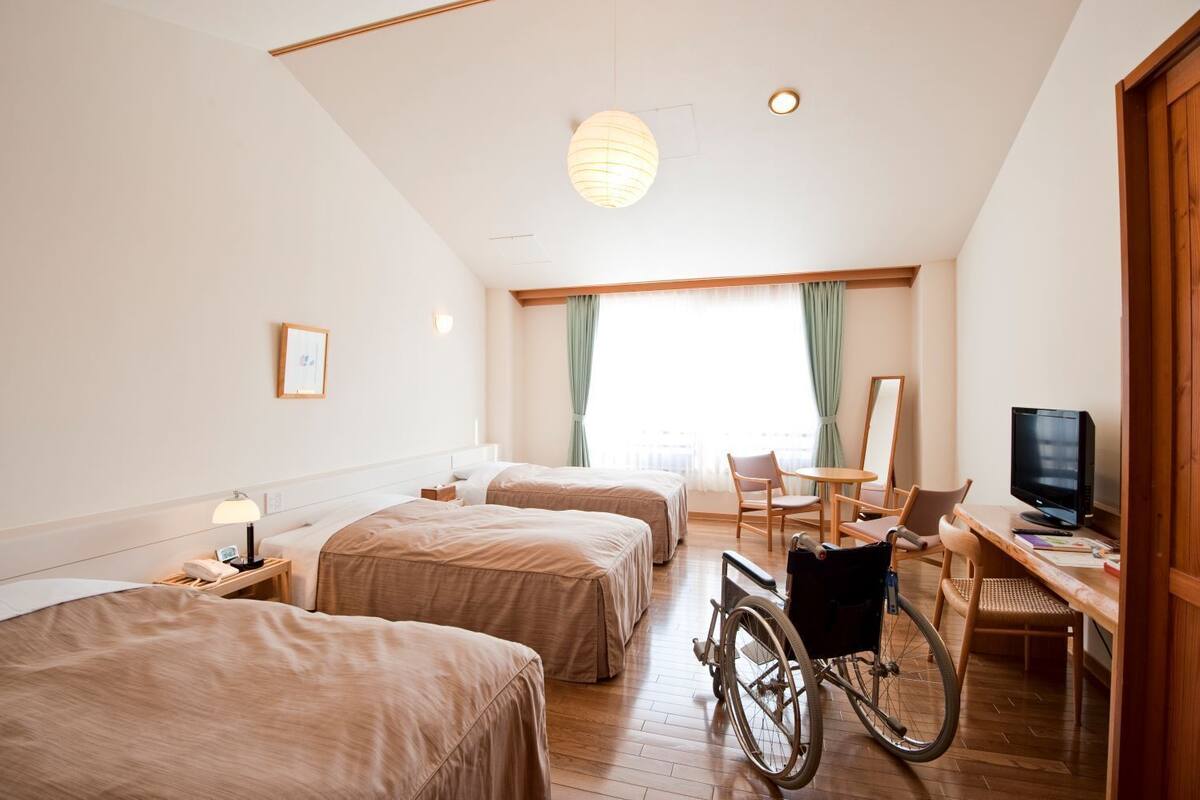 Quarto de hotel com duas camas, uma cadeira de rodas, uma mesa com computador e uma janela grande com cortinas.