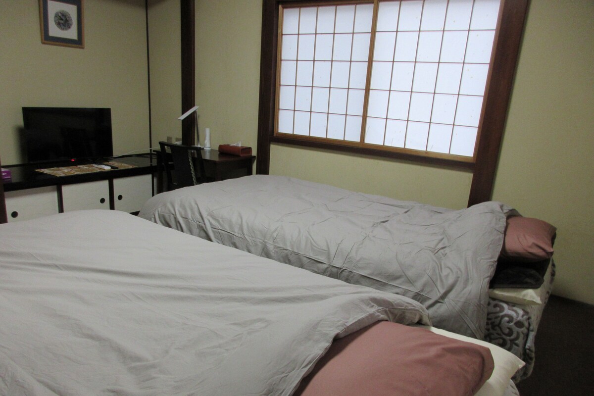 Quarto com duas camas, uma escrivaninha com computador e janela com padrão quadriculado.