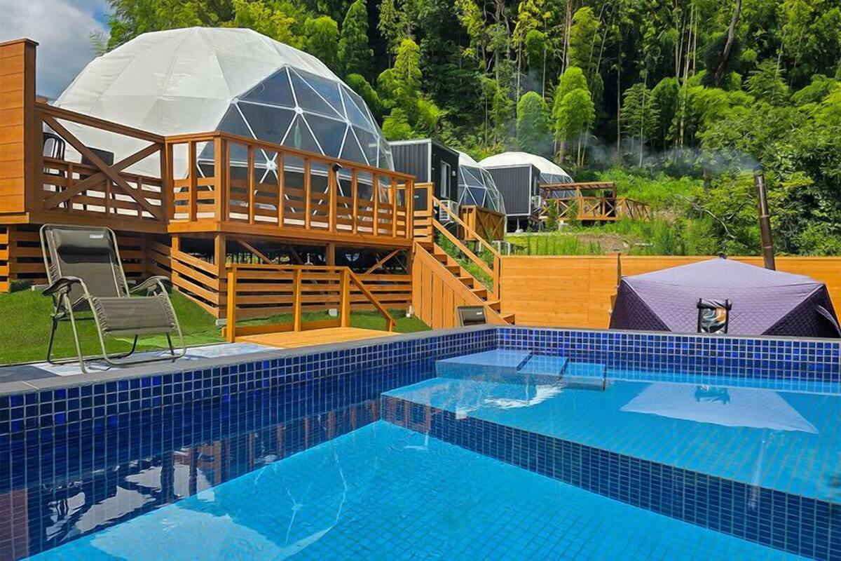 Uma piscina com deck de madeira, estrutura em cúpula de vidro e vista para uma área arborizada.