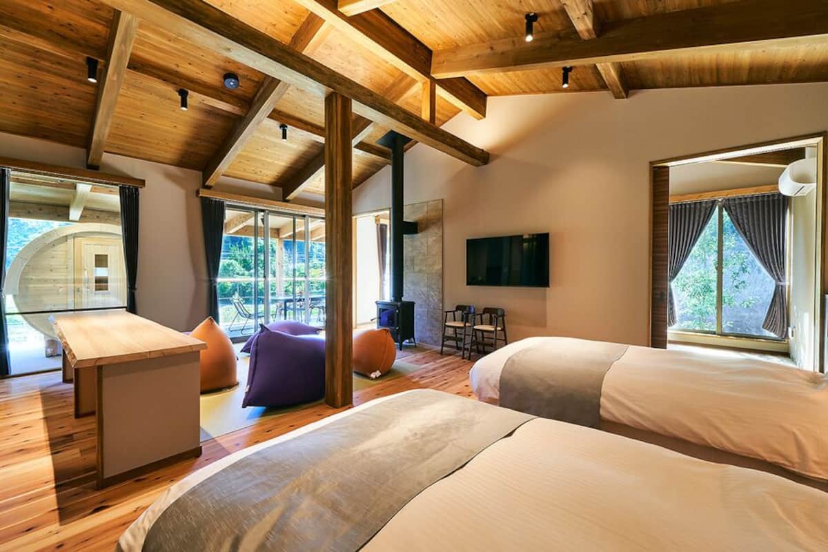Quarto moderno com vigas de madeira, duas camas, uma TV e vista para o exterior.