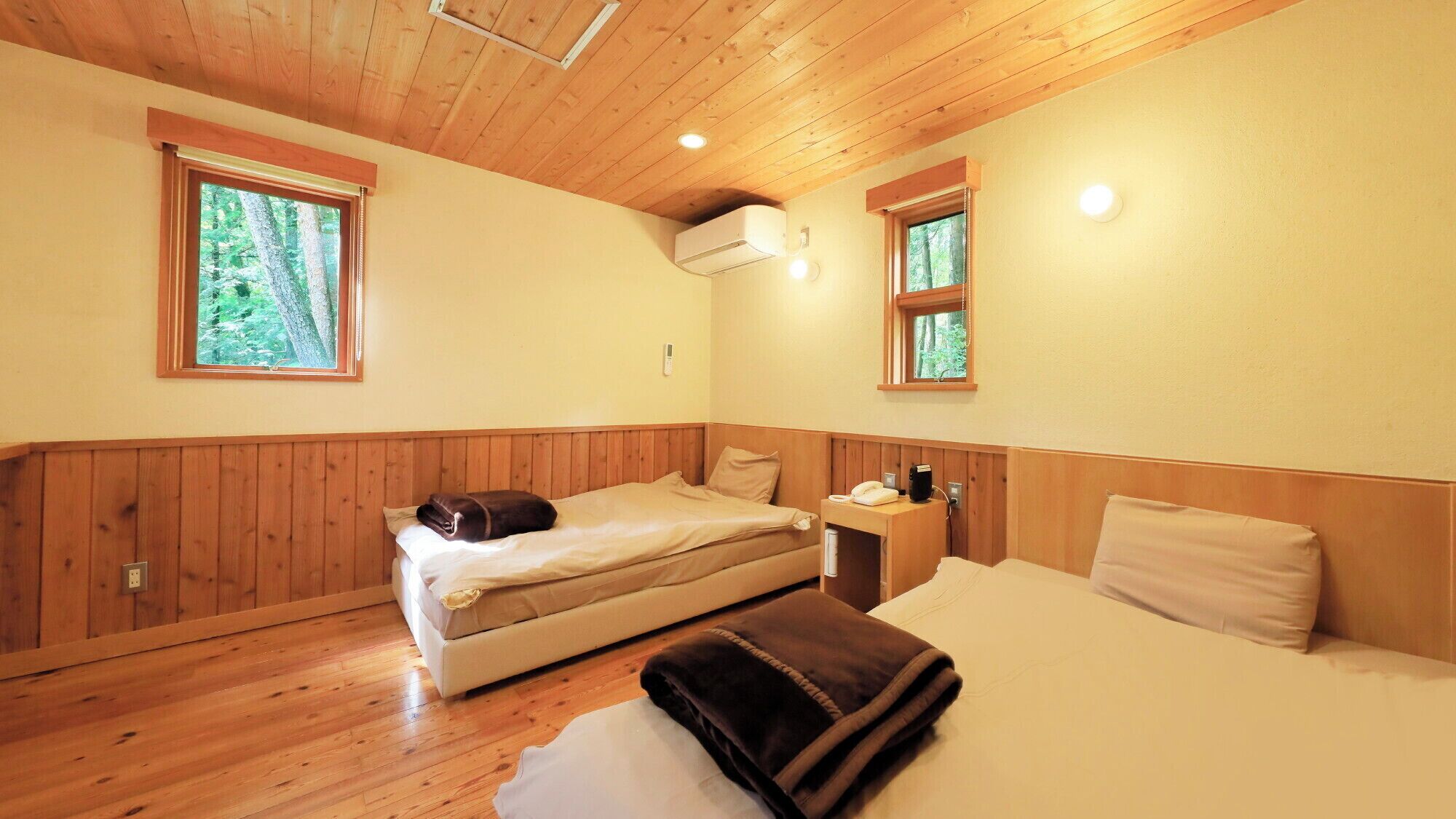 Quarto com teto de madeira, duas janelas com vista para o verde, uma cama com cobertor, mesa de cabeceira com telefone e unidade de ar condicionado.