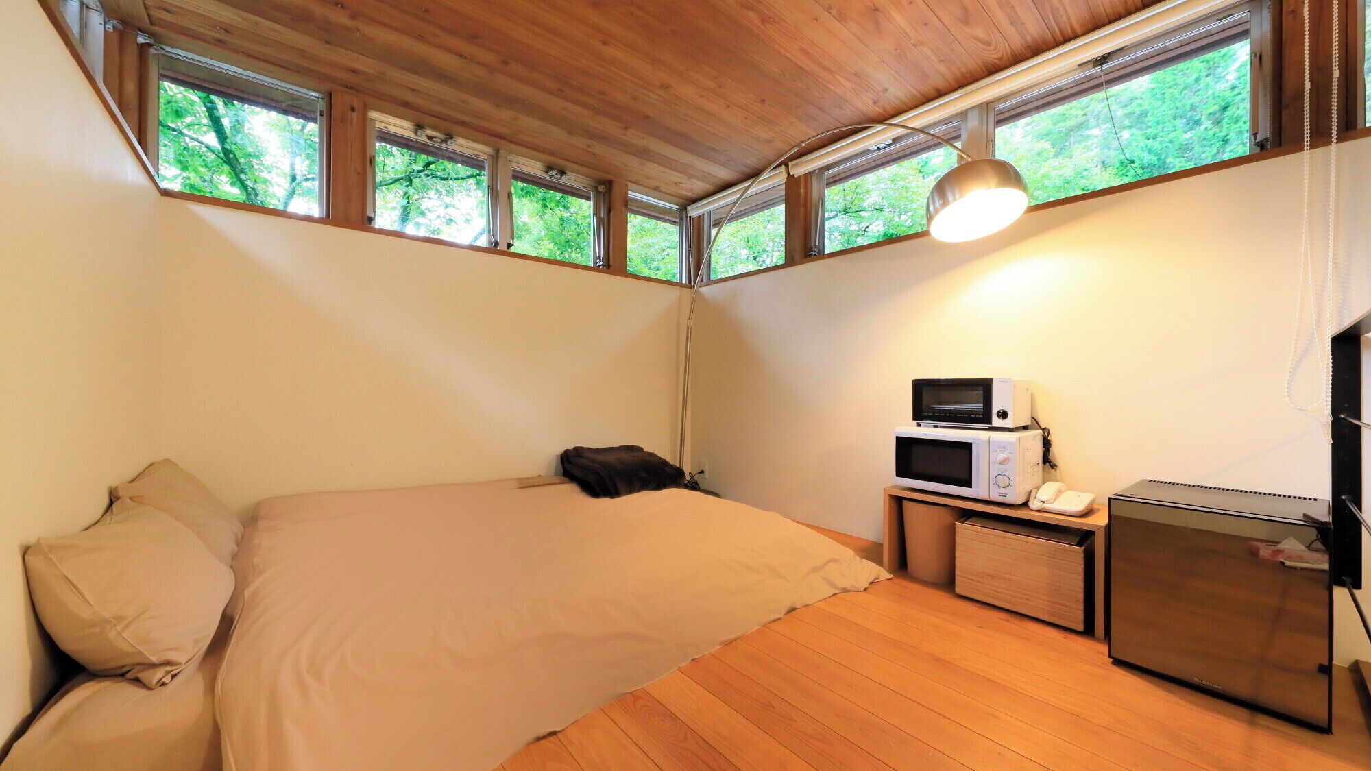 Quarto com cama, mesa de cabeceira com micro-ondas e telefone, escrivaninha com computador e janelas amplas.