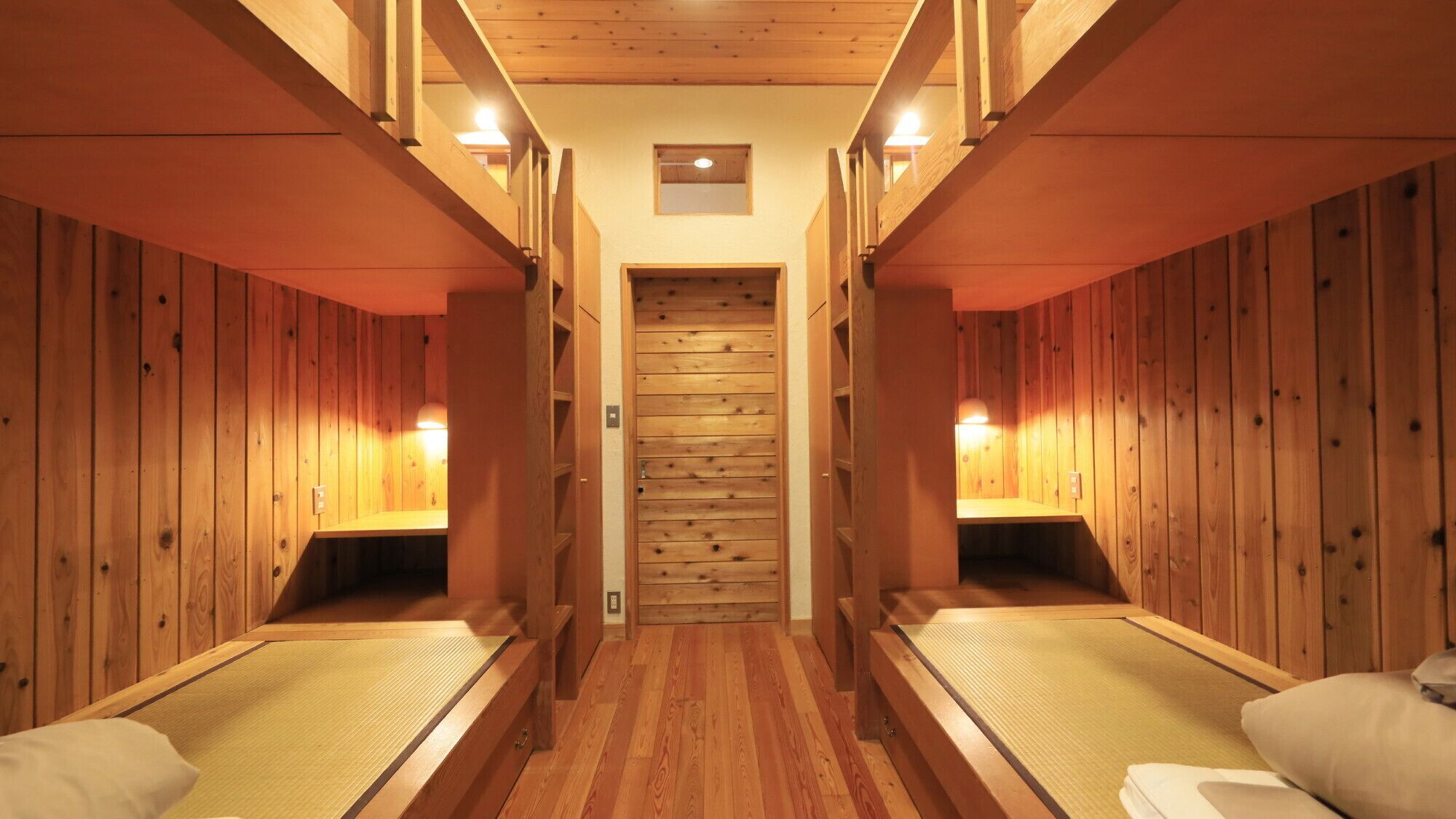 Fileira de cabanas de madeira com camas individuais, porta central e luminárias fixadas na parede.