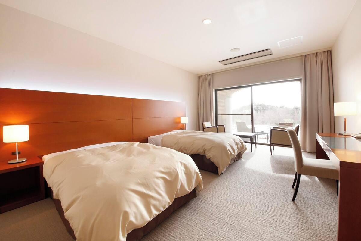 Quarto de hotel com duas camas, uma escrivaninha, uma cadeira e uma janela grande.
