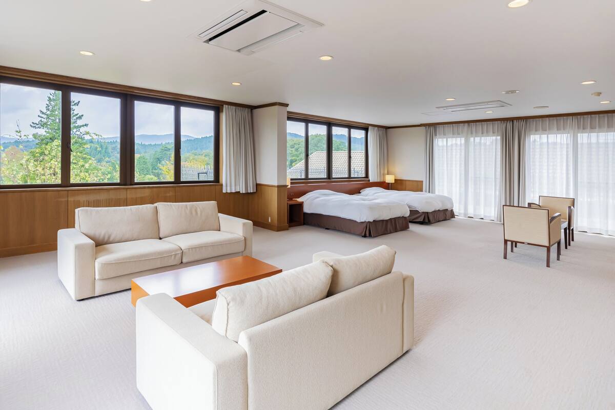 Quarto de hotel moderno com uma cama grande, dois sofás, uma mesinha e amplas janelas com vista para as montanhas.