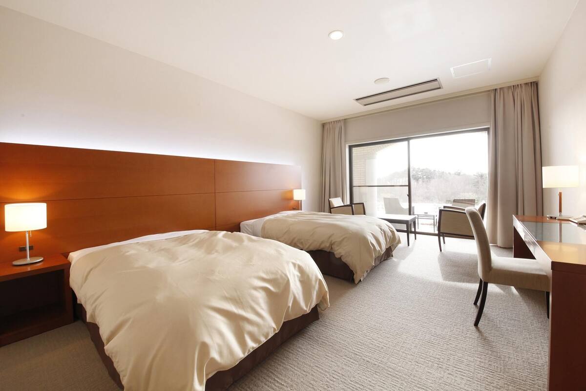Quarto de hotel com duas camas, uma escrivaninha, uma cadeira e uma janela grande.
