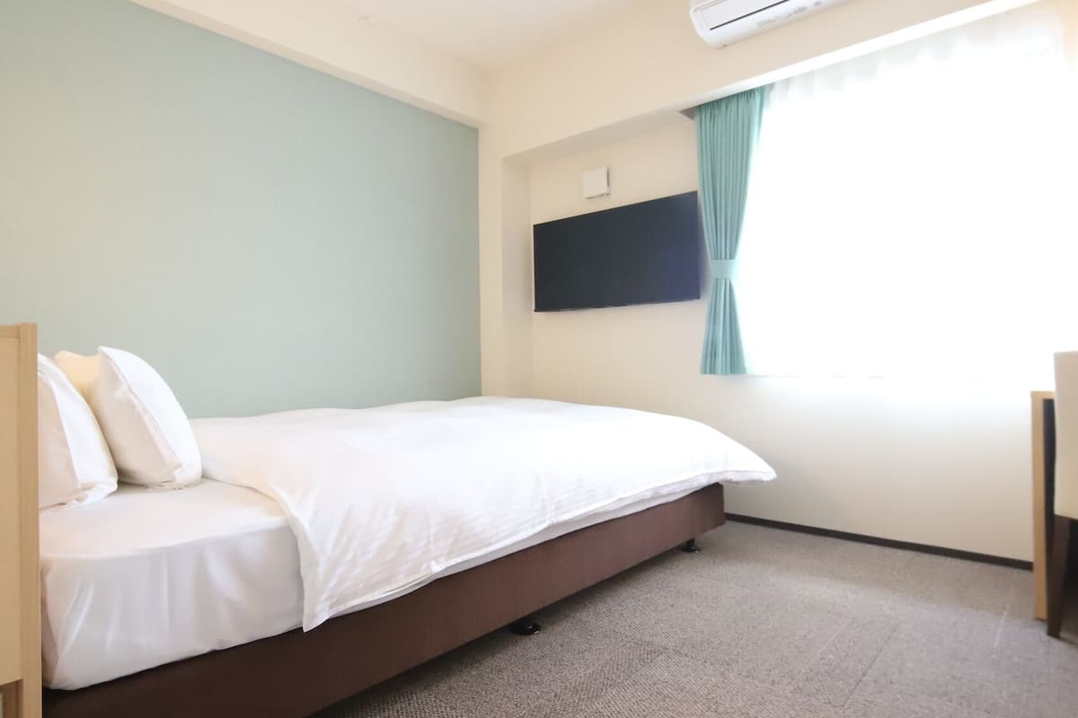 Quarto de hotel com uma cama, televisão de tela plana na parede, janela com cortinas e uma pequena mesa com cadeira.