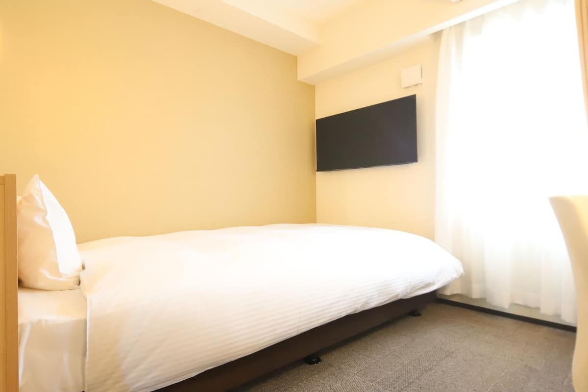 Uma cama de solteiro com cabeceira, mesa de cabeceira, televisão de tela plana na parede e uma janela com cortinas.