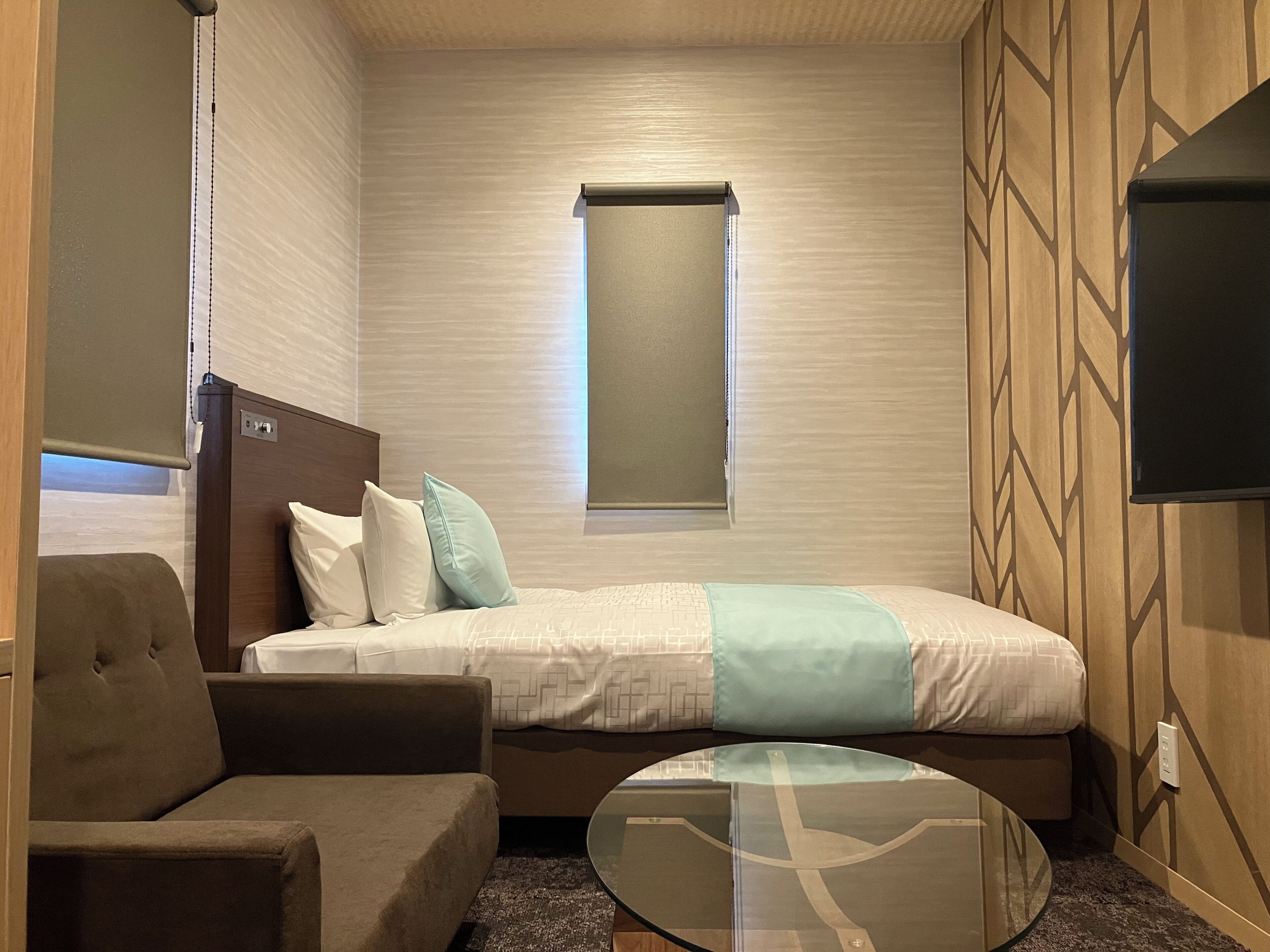 Quarto de hotel compacto com cama, um pequeno sofá, mesa de centro de vidro, televisão e janela com persianas.