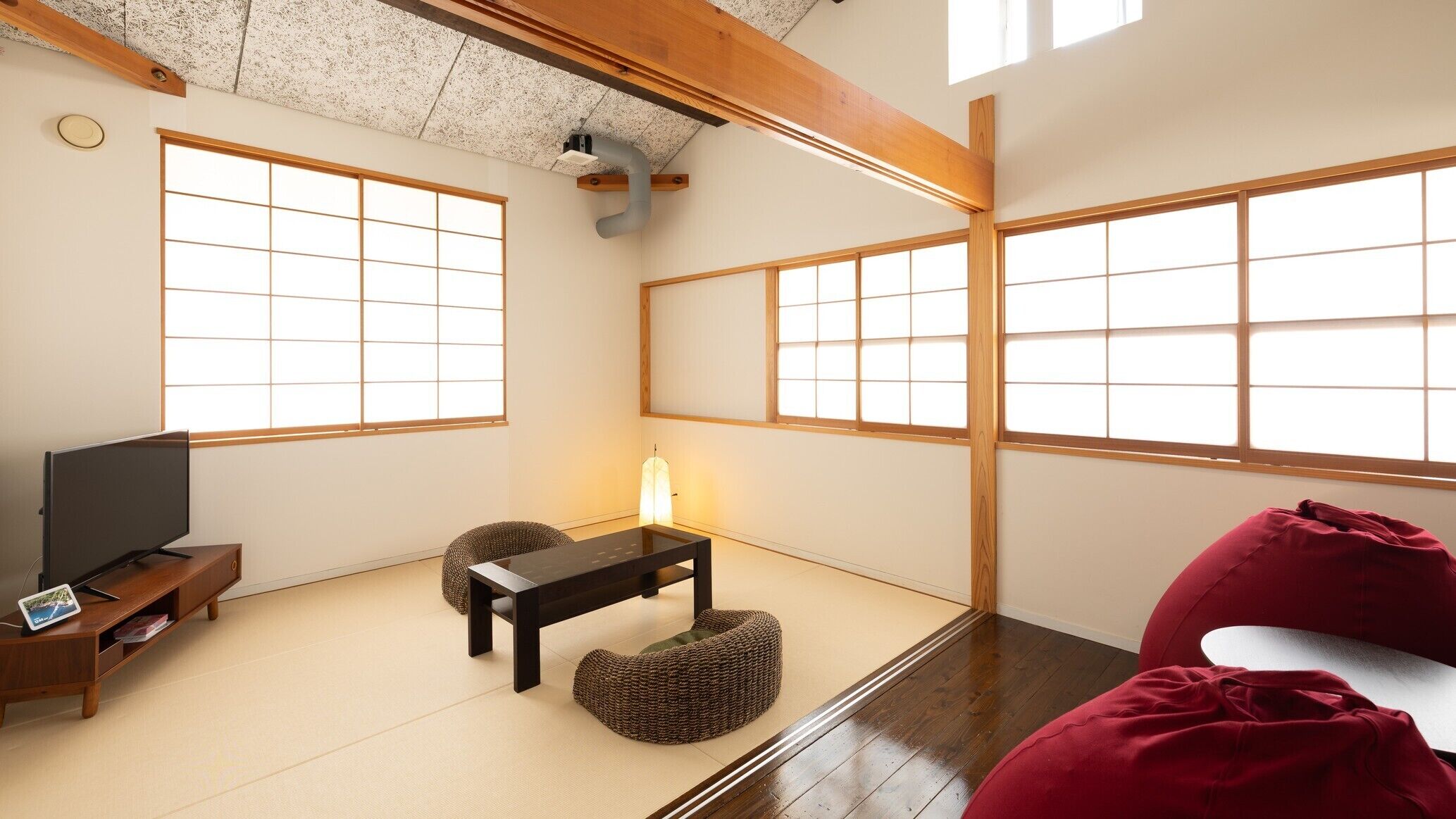 Quarto japonês tradicional com tatames, portas deslizantes shoji, uma televisão, uma mesinha e um pufe vermelho.