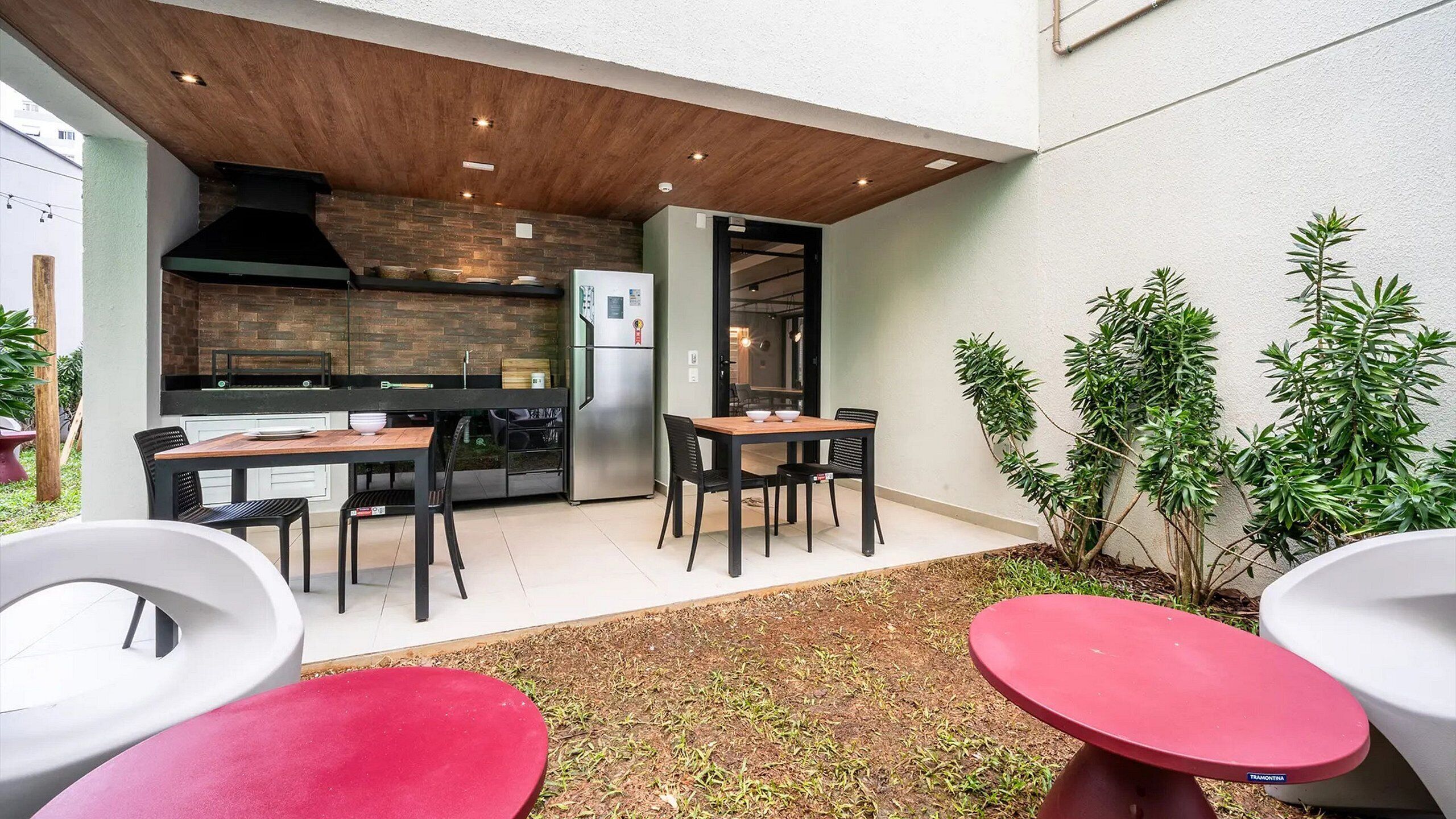 Cozinha moderna com parede de tijolos, eletrodomésticos de aço inoxidável e teto de madeira. Há um espaço de jantar com mesa e cadeiras, e um pequeno jardim com plantas e uma mesa vermelha.