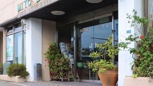 Exterior de um hotel com a placa 'MILTON DAANCHI HOTEL'. Há plantas em vasos na frente da entrada, e uma janela com reflexo visível.