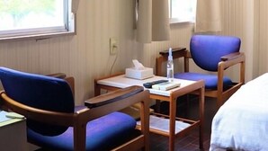 Quarto de hotel com uma cama, duas cadeiras azuis, uma mesinha com um estojo de lenços, um controle remoto e uma garrafa.