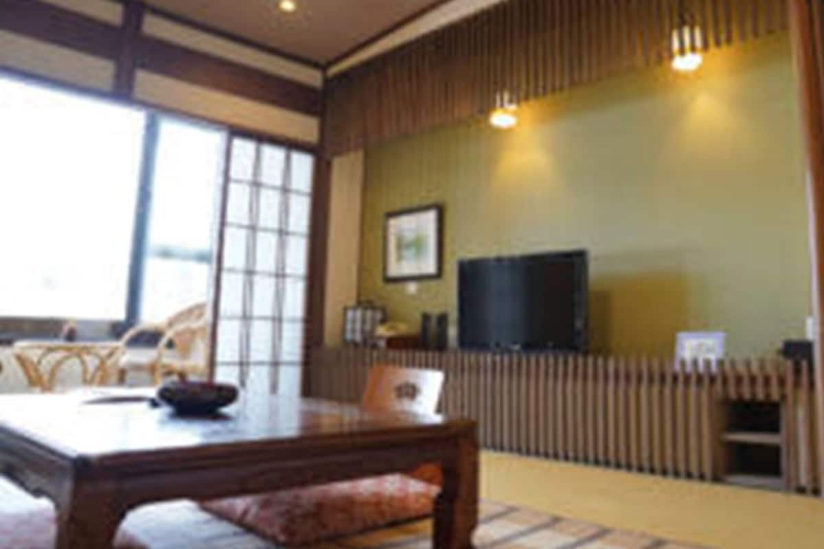 Uma sala de estar tradicional japonesa, com tatames no chão, uma mesa de madeira baixa e uma televisão presa na parede.