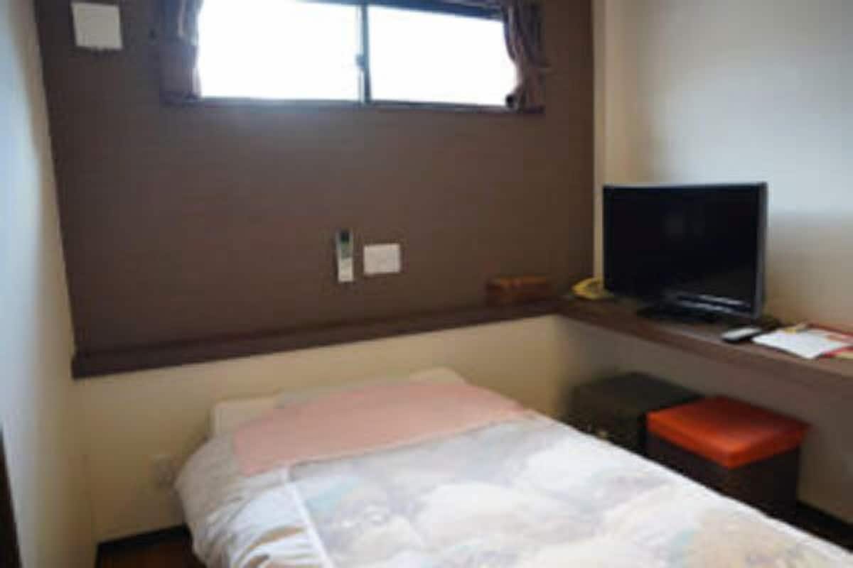 Um quarto pequeno com uma cama, uma escrivaninha com um computador, uma janela e uma televisão.