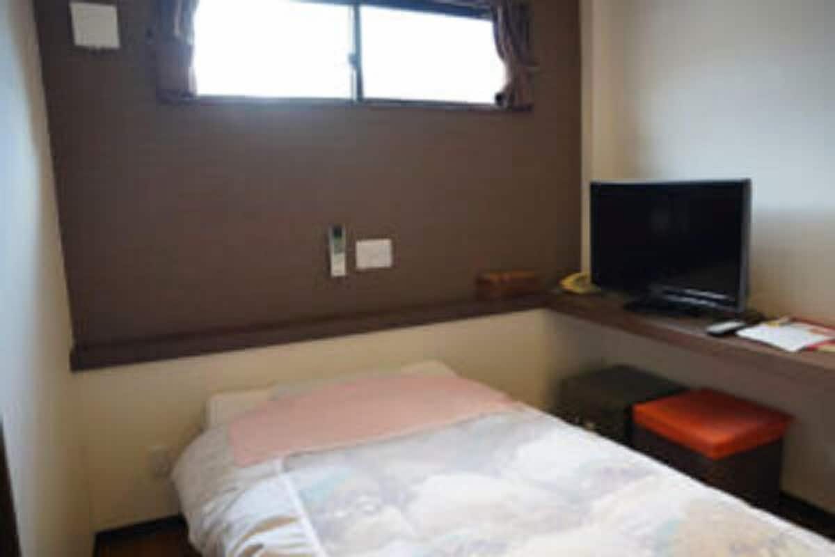 Um quarto pequeno com uma cama, uma escrivaninha com um computador, uma televisão e uma janela.