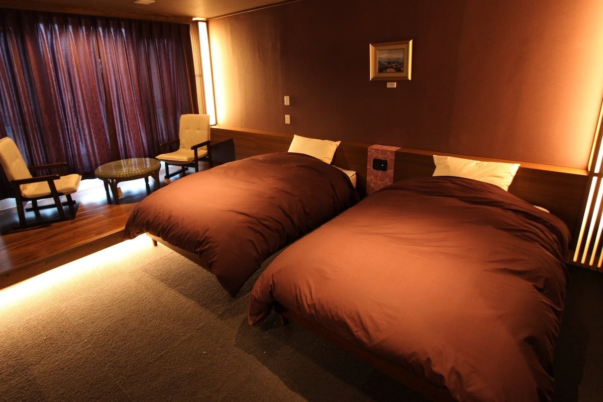 Quarto de hotel com duas camas, uma mesinha, cadeiras e uma janela grande com cortinas.