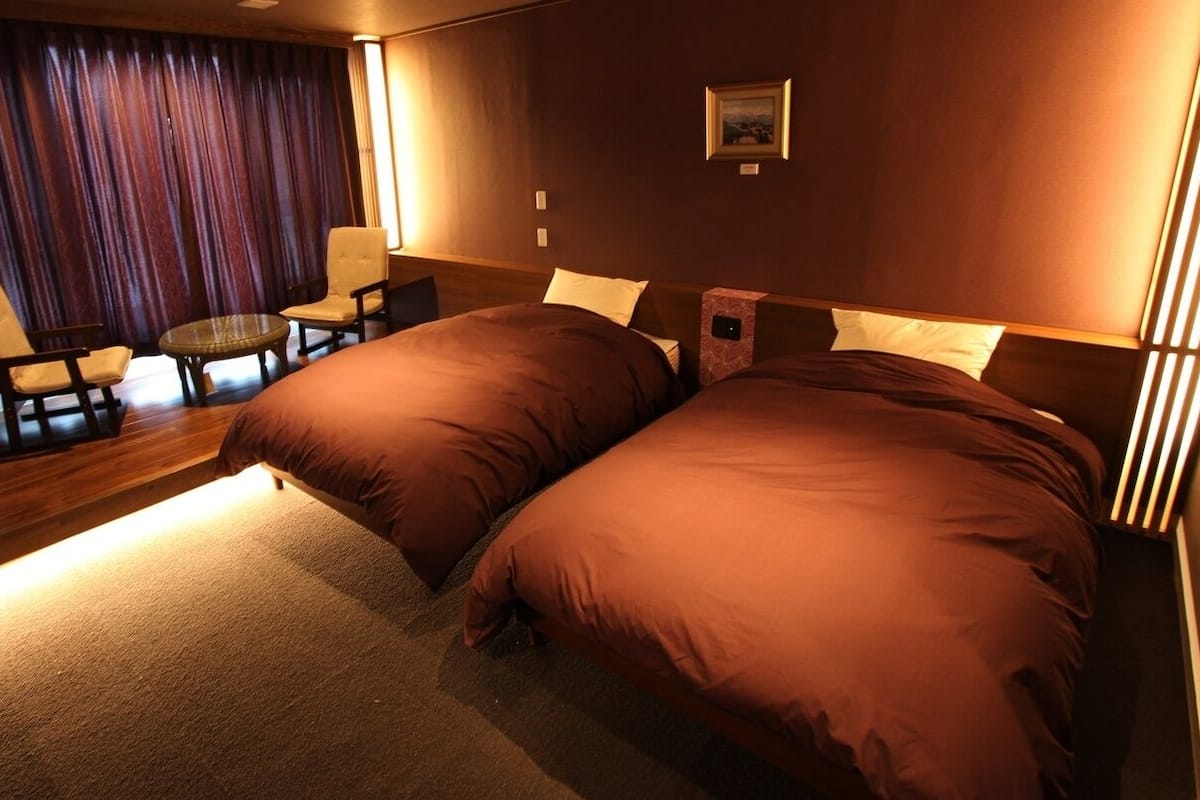 Quarto de hotel com cama, duas cadeiras, uma mesinha e cortina.