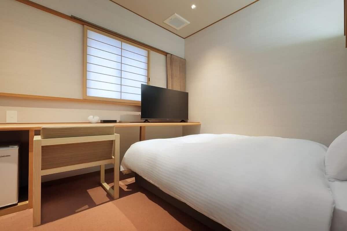 Quarto de hotel com cama, escrivaninha e televisão, e janela com persianas.