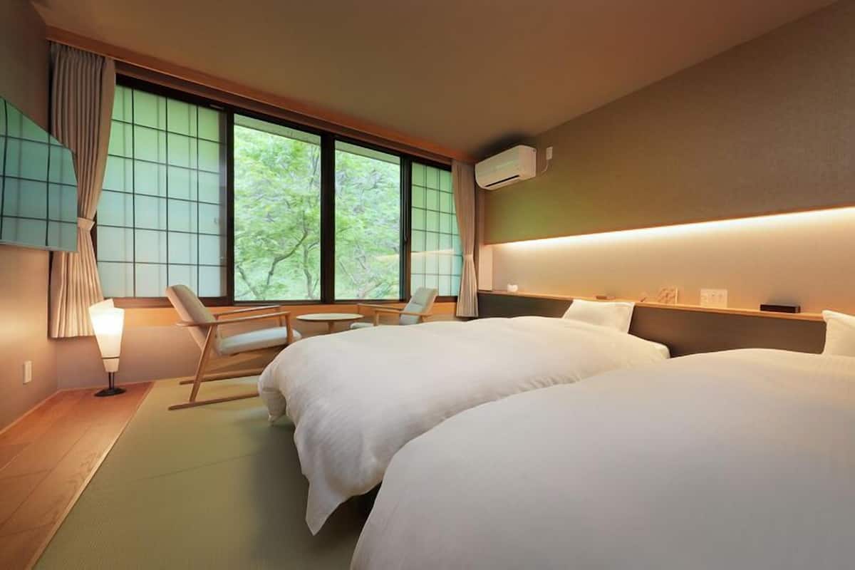 Quarto de hotel com duas camas, área de estar com uma cadeira e mesa, e uma janela grande com cortinas.