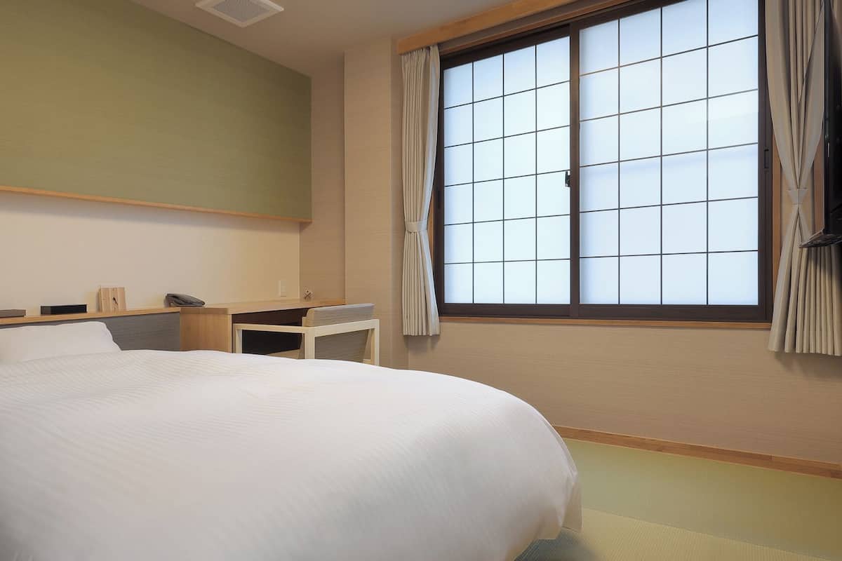 Quarto de hotel com uma cama grande, uma pequena escrivaninha, uma cadeira, uma janela com cortinas e uma estante fixada na parede.