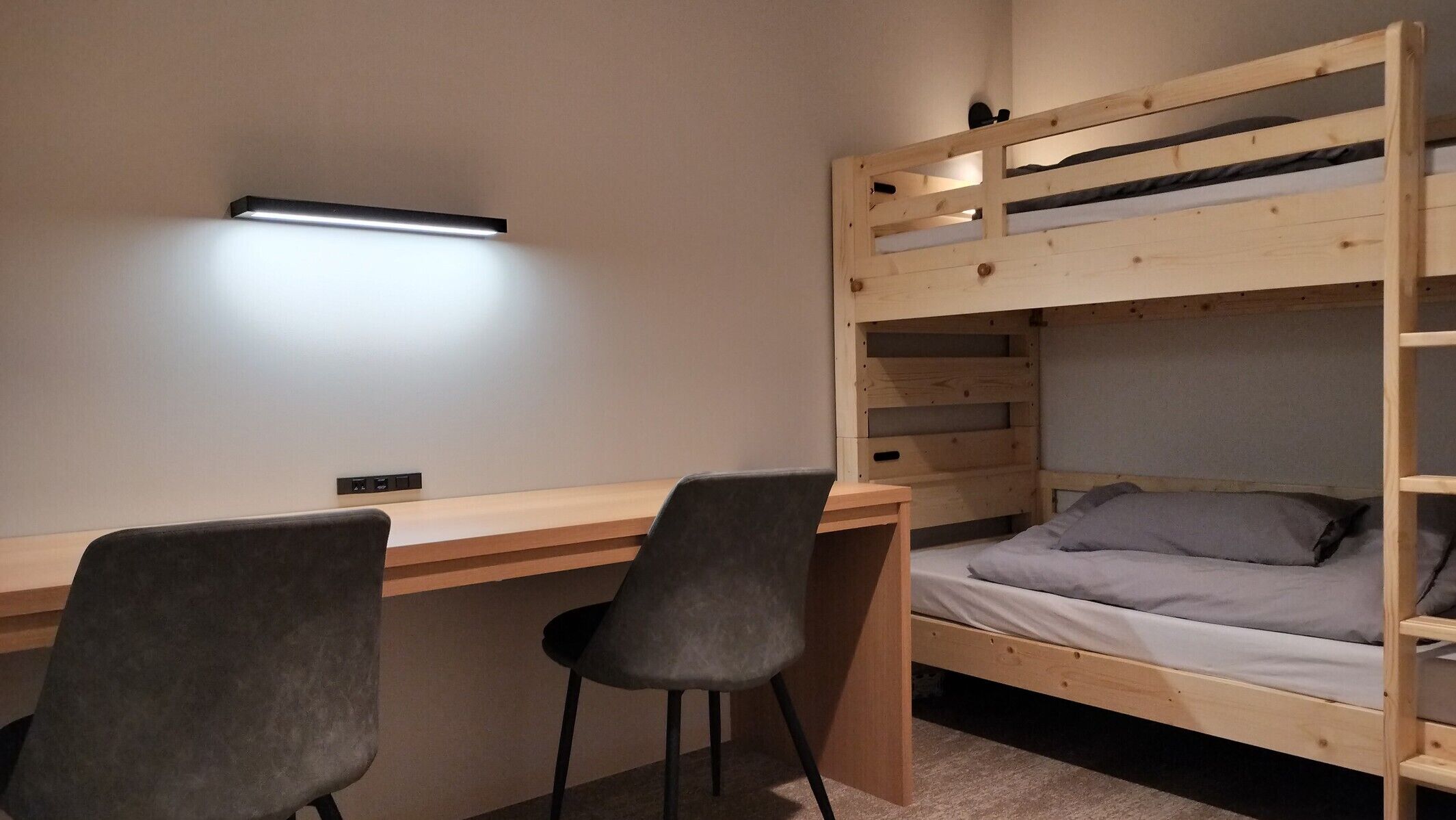 Uma cama beliche com escrivaninha, duas cadeiras, mesa de cabeceira e luminária fixa na parede.