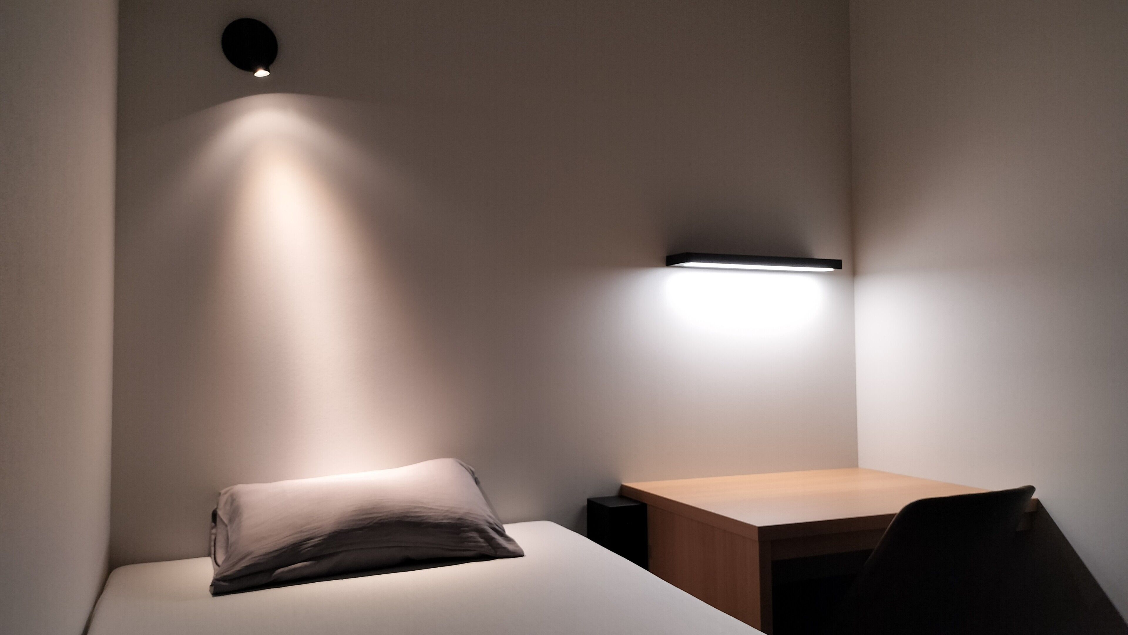 Quarto pequeno e minimalista com uma cama, uma escrivaninha e uma luminária na parede.