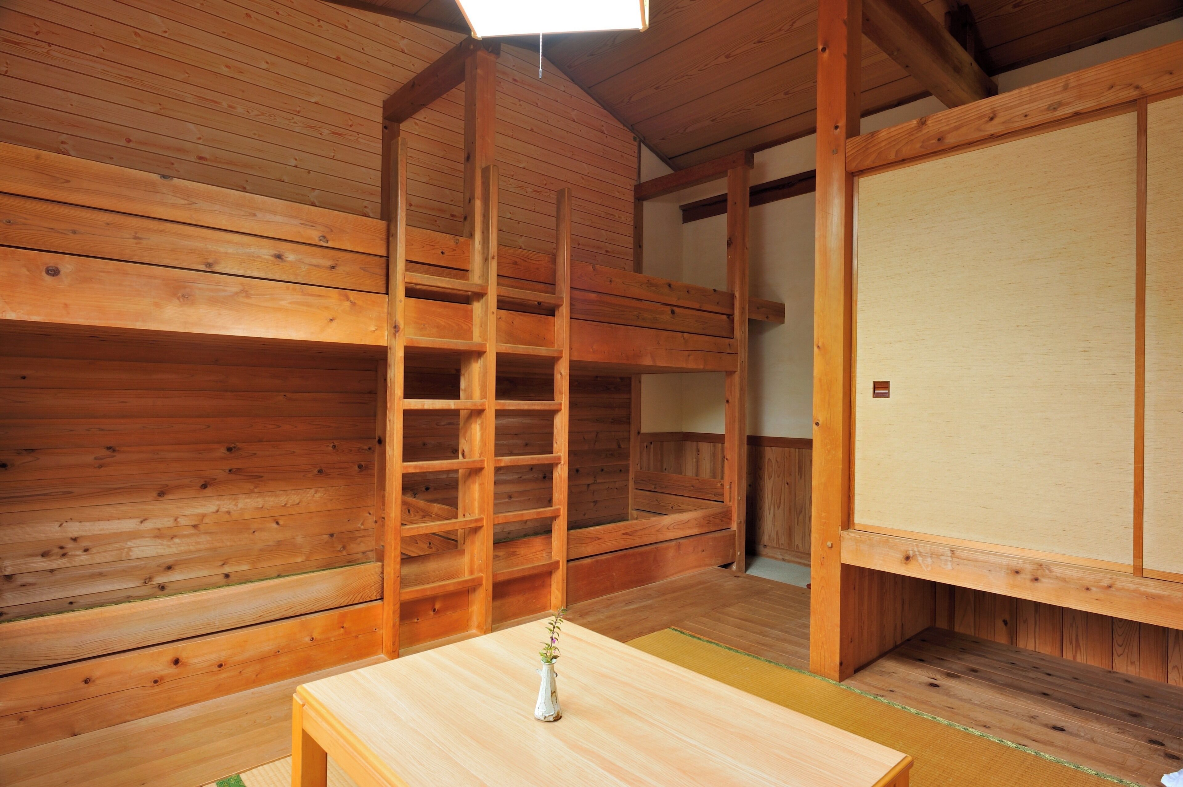 Interior em madeira com beliches, uma escada, uma mesinha com um vaso e uma porta deslizante.