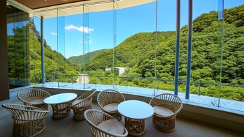 Varanda com mobiliário de vime e parede de vidro, com vista para um vale verde e um edifício.
