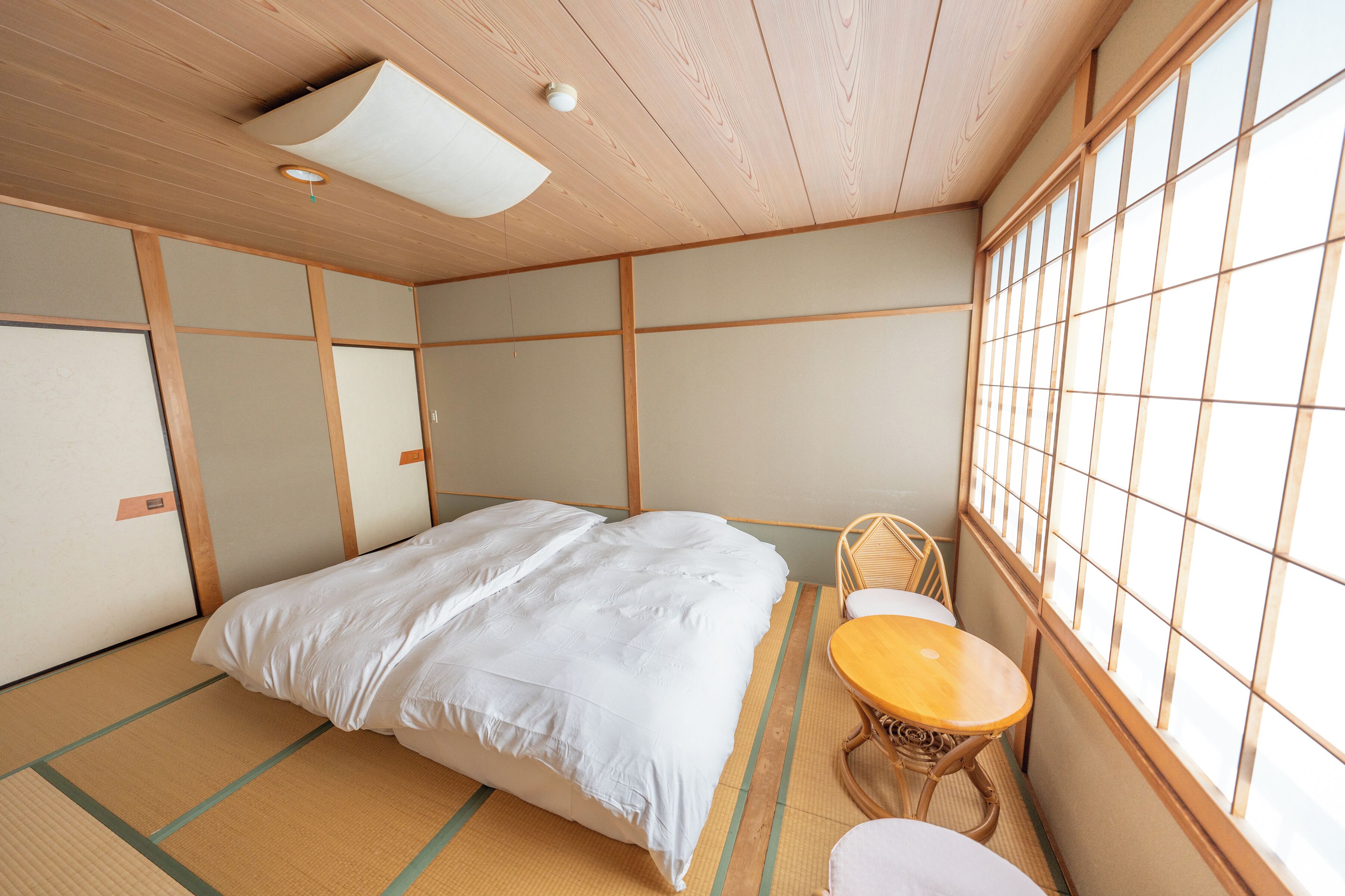 Um quarto japonês tradicional com piso de tatame, uma cama com roupa de cama branca, uma pequena mesa de madeira e uma janela com desenhos em grade.