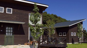 Uma casa de madeira de dois andares com varanda frontal, uma porta verde e um pequeno jardim com vasos de plantas.