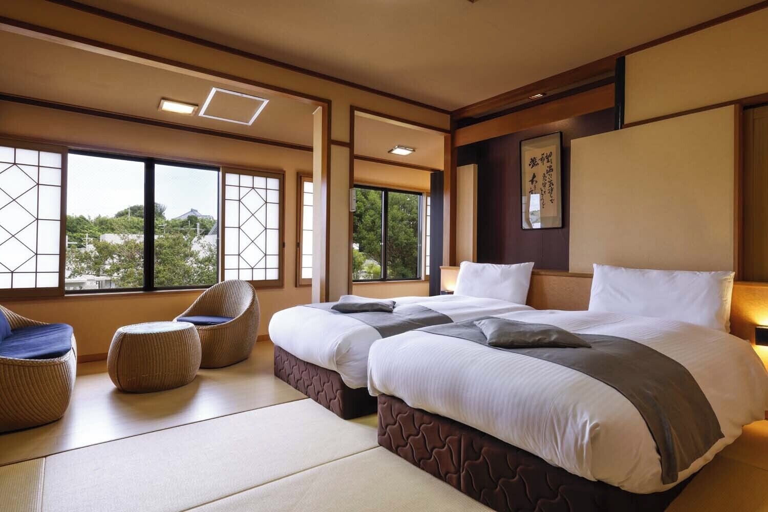 Quarto de hotel com piso de tatame, uma cama grande com cabeceira, duas áreas de estar com almofadas e amplas janelas com vista para o verde.