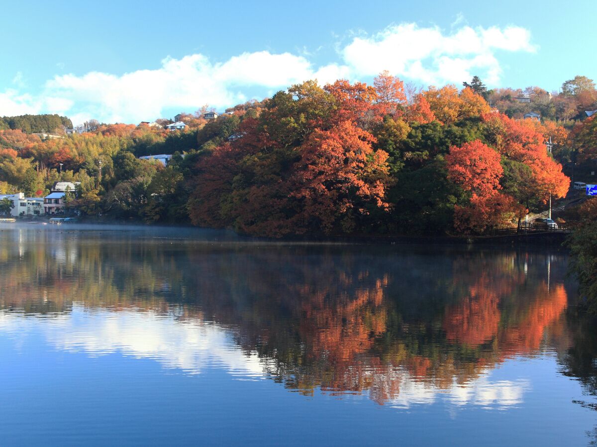 Um lago calmo com folhagem outonal refletida na água, rodeado por colinas e edifícios.